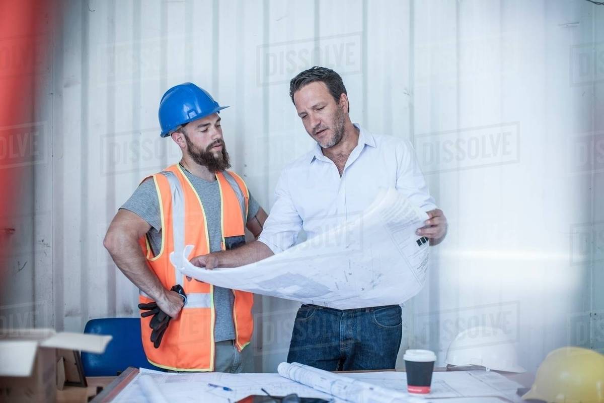 Construction foreman looking over blueprint with worker at desk in ...