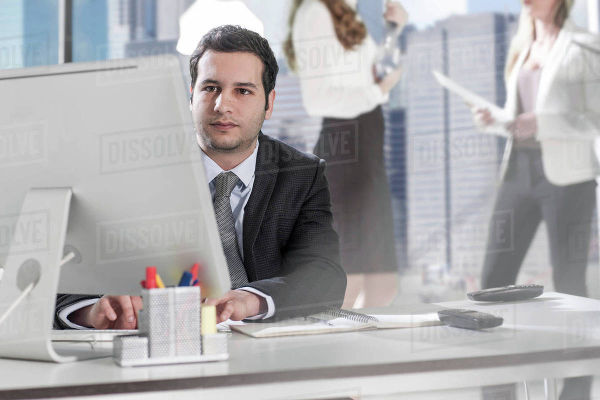 Businessman using computer in office, colleagues chatting in background ...