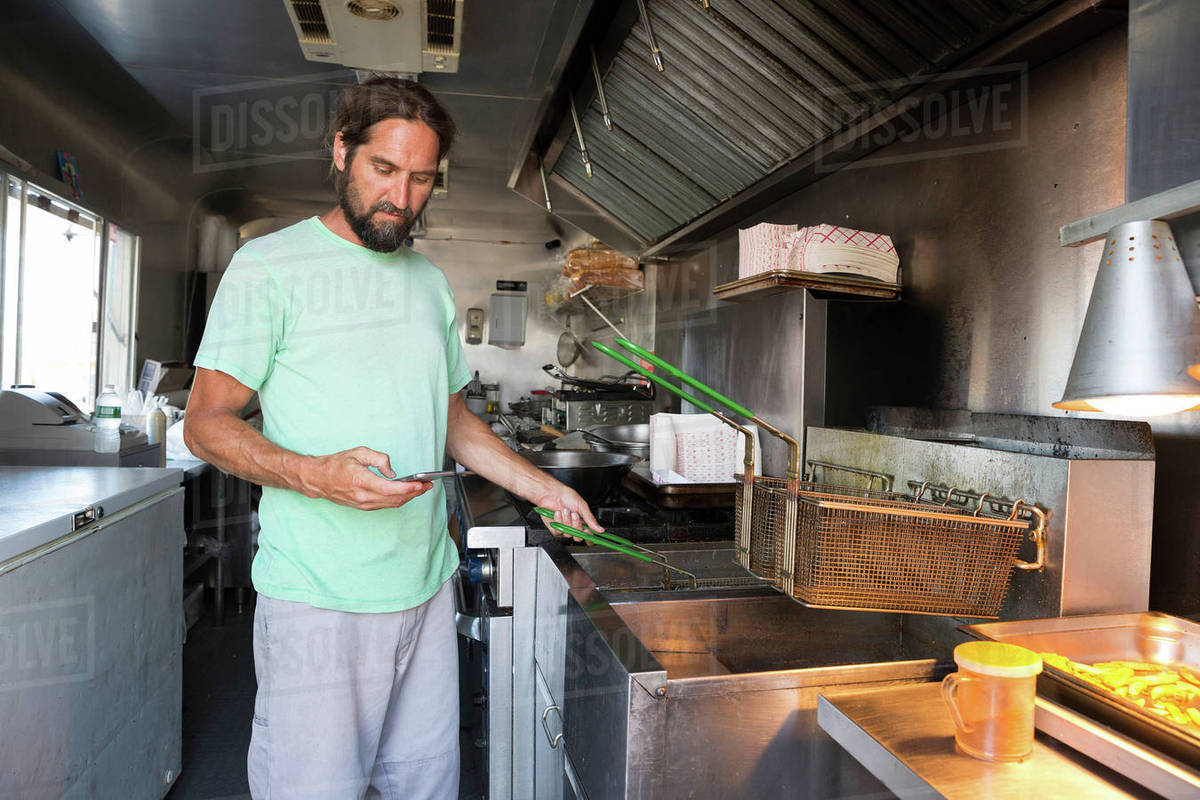 Man cooking food in fast food trailer whilst looking at smartphone ...