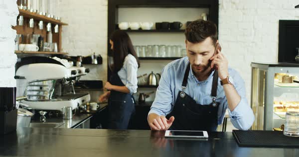 Young handsome waiter talking on the phone and scrolling on the tablet ...