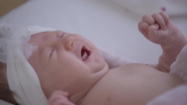 Side view crying newborn baby girl in white hair hoop lying on bed ...