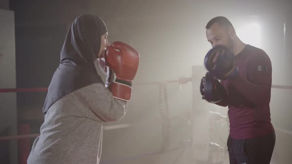 Side view of muslim female boxer punching training gloves as coach ...