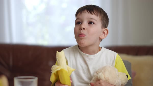 Close-up. A child looks at the camera and bites into a peeled banana. A ...
