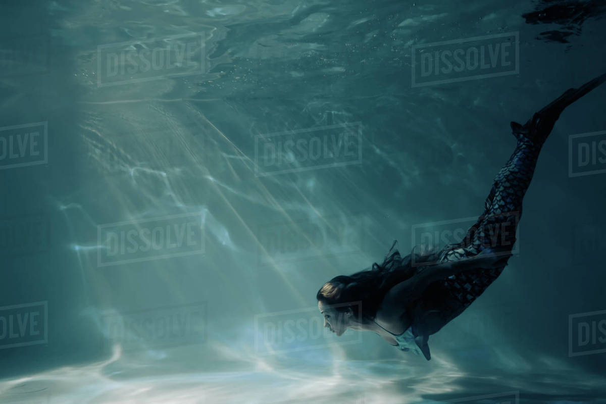 Beautiful young woman with mermaid tail swims and dives under sunlit ...