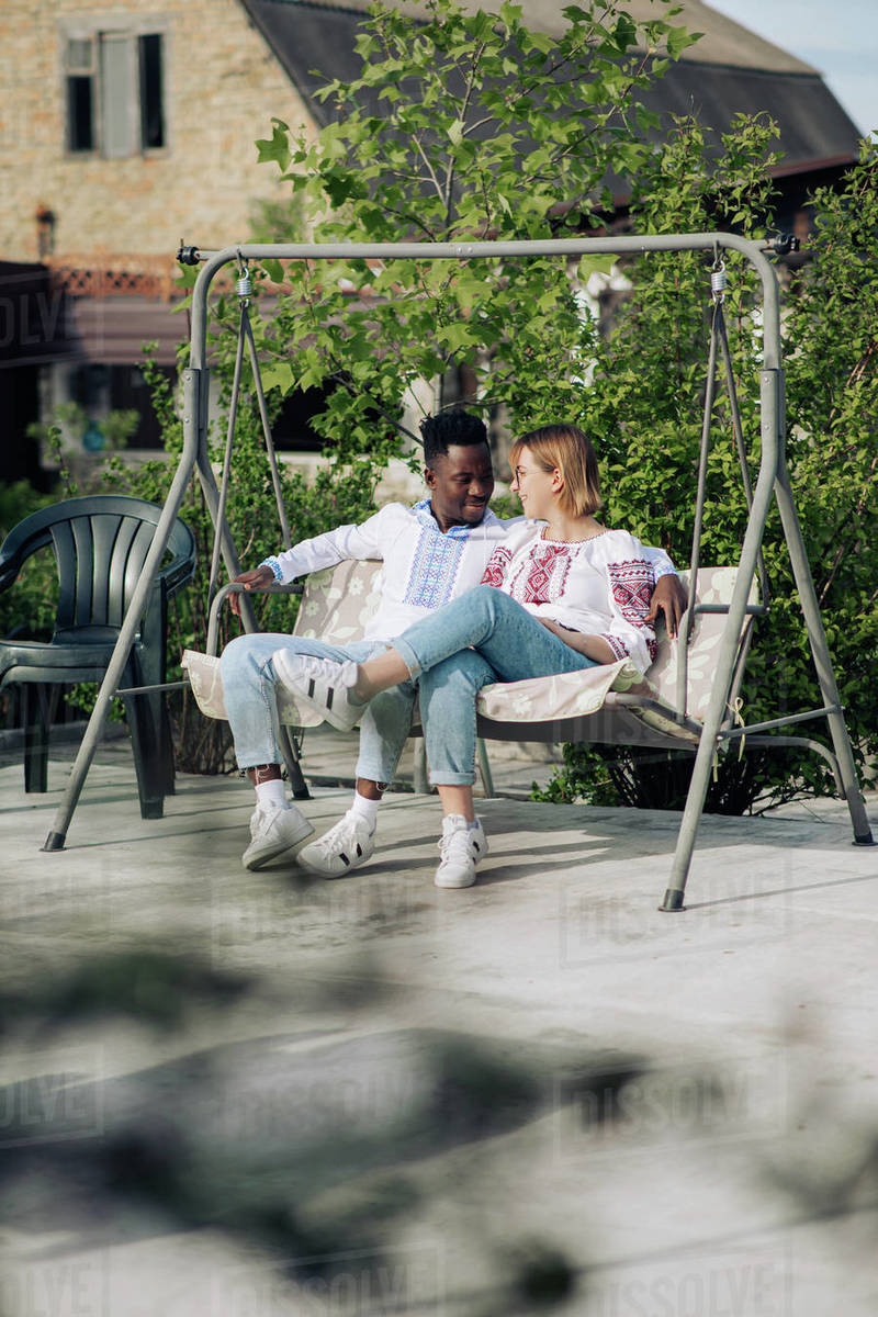 Interracial couple sits on bench swing in garden dressed in Ukrainian ...