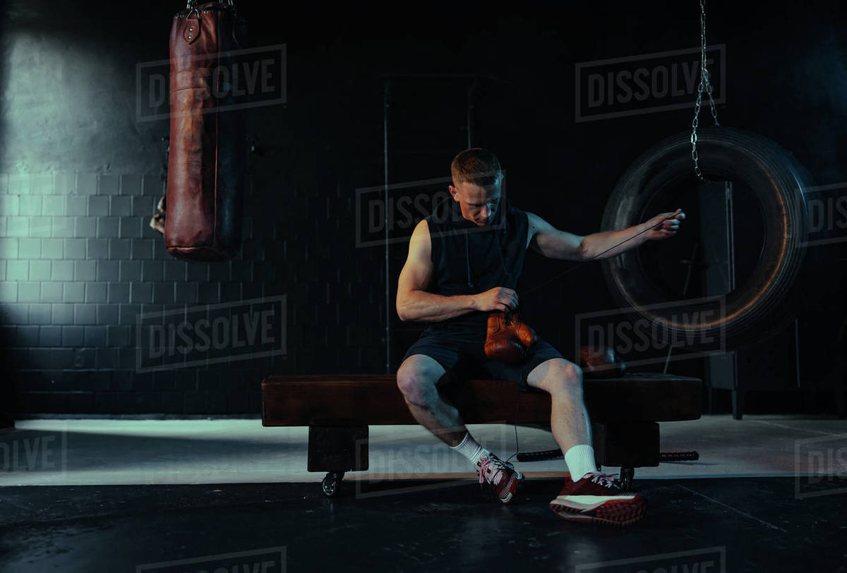 Male boxer sitting on bench in dark gym and putting on boxing gloves ...