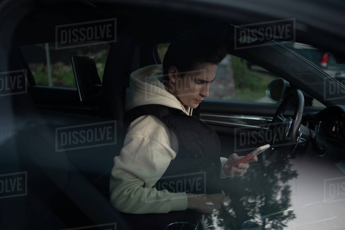Side view of a young man using smartphone while driving a car on street ...