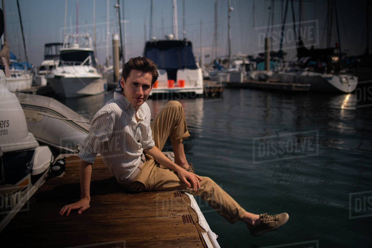 Portrait of a young man sitting on wooden pier at harbor - Royalty-free ...