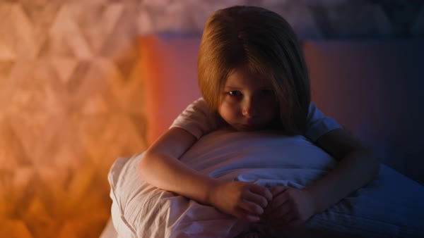 Sad little daughter with dimly lit face sits leaning on big soft pillow ...