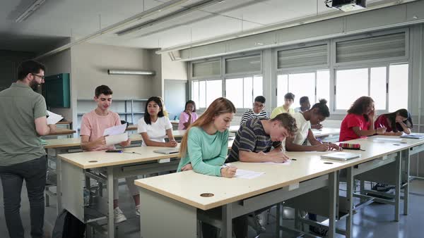 Young teacher giving test sheets to students in class. Multi-ethnic ...