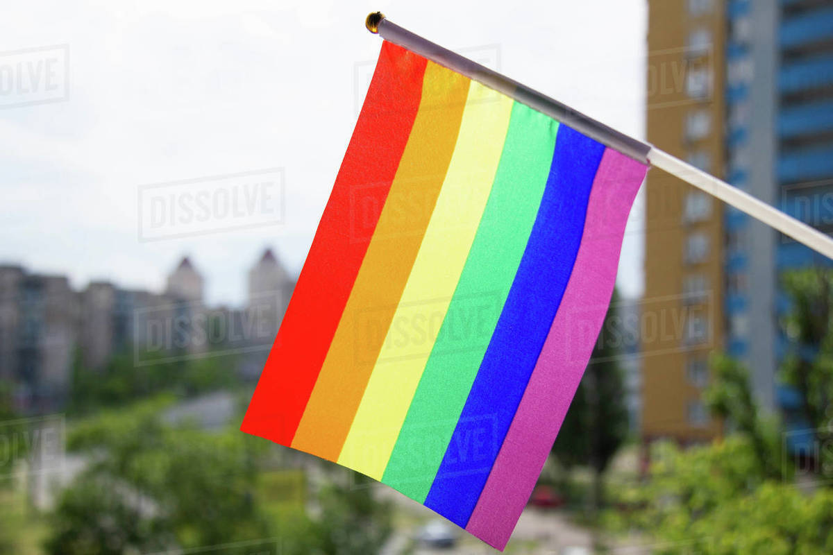 Rainbow LGBT flag set against blue sky, green trees, city street and ...