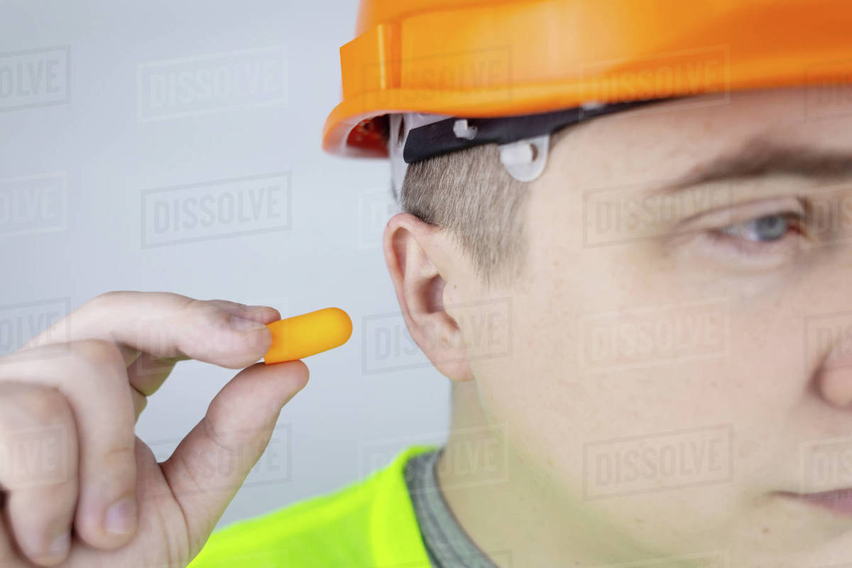 A worker in a hard hat and green vest puts on ear plugs. Industrial ...