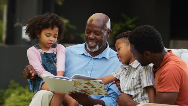 Multi-generational family sitting in garden at home having fun reading ...