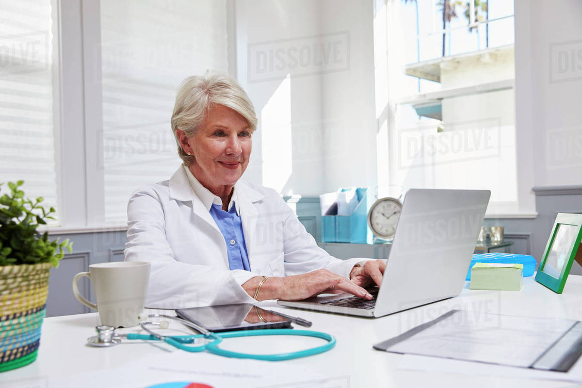 Female Doctor Sitting At Desk Working At Laptop In Office - Royalty ...