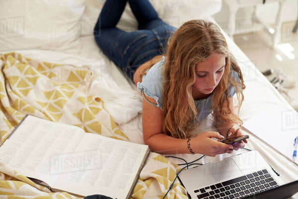 Girl lying on bed using smartphone and laptop, high angle - Royalty ...