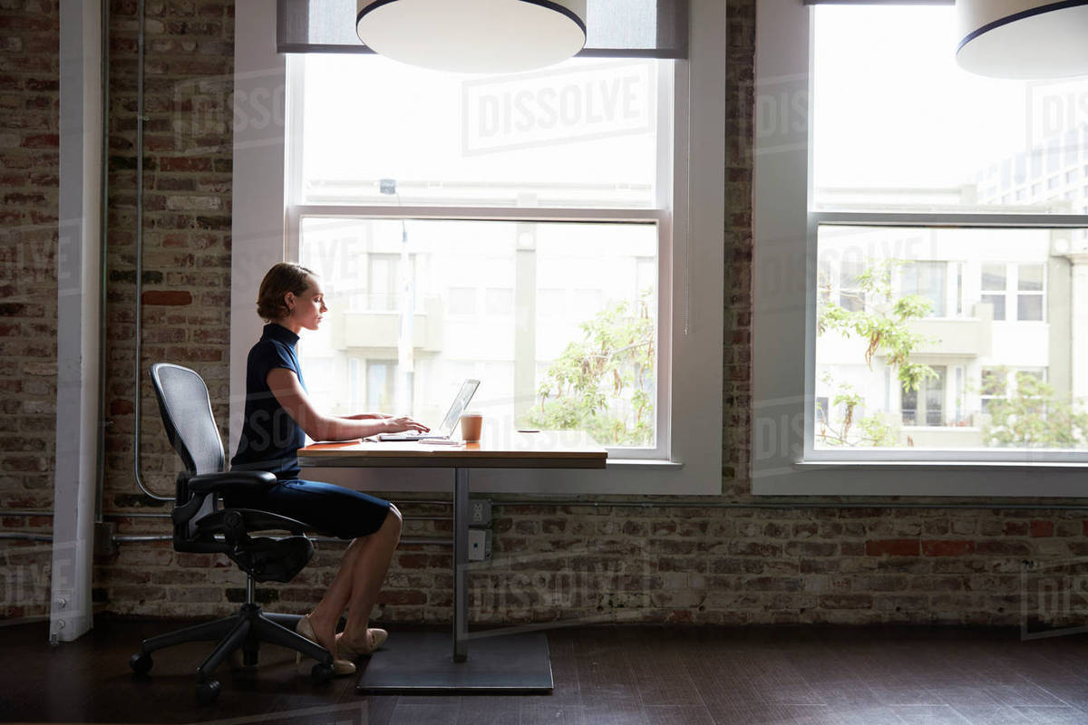 Businesswoman sitting at desk by window working on laptop - Stock Photo ...