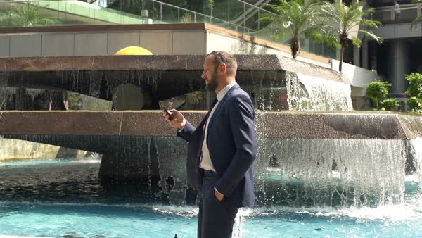 Businessman texting on smartphone walking near fountain in city, super ...