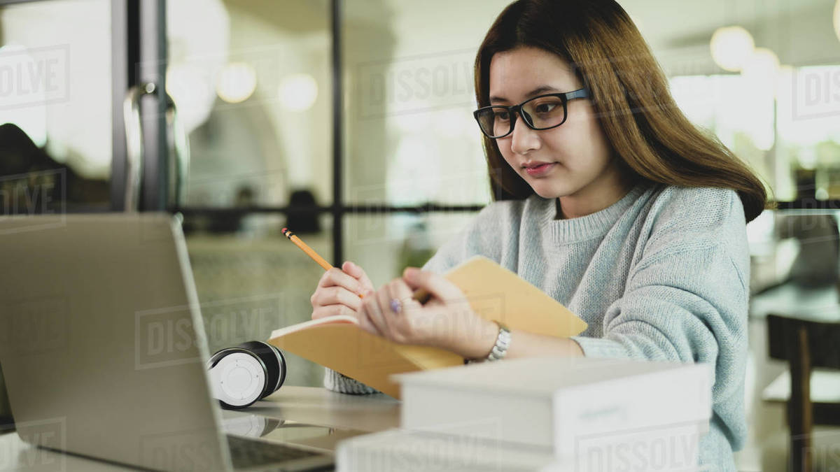 Asian female student wearing glasses looking at laptop and taking notes ...