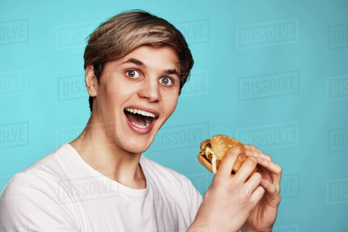 Very hungry young man holding tasty hamburger isolated over blue background. copy space - Stock ...