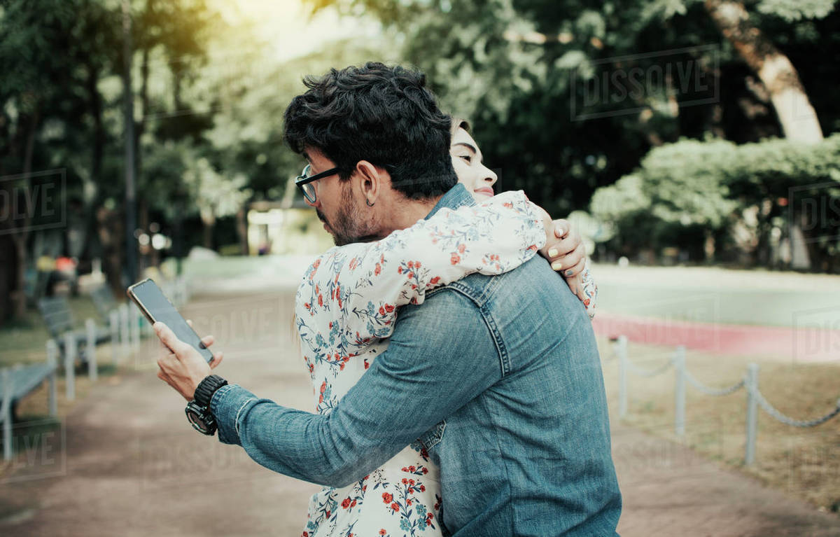 Unfaithful man hugging his girlfriend and looking at the cell phone ...
