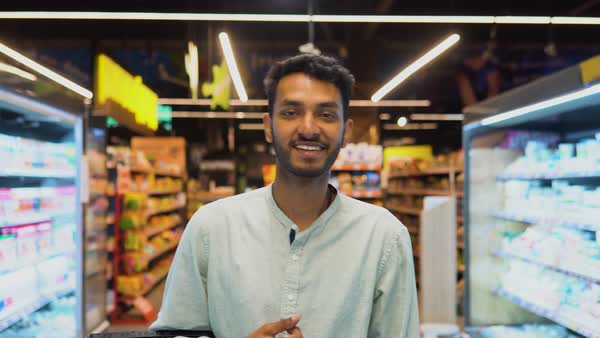 Smiling indian man walking in aisle with shopping basket at supermarket ...