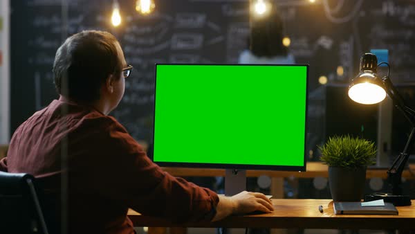 Office employee at his desktop works on a mock-up green screen personal ...