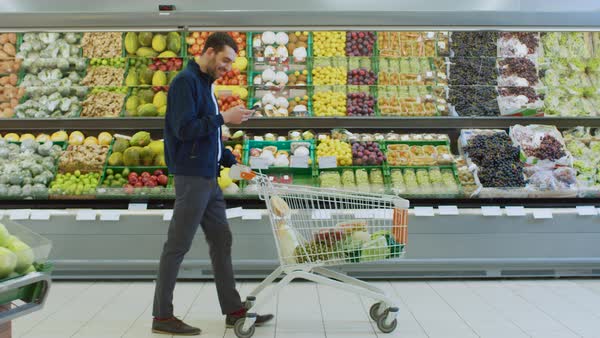 At the supermarket: handsome man with smartphone, pushes shopping cart ...
