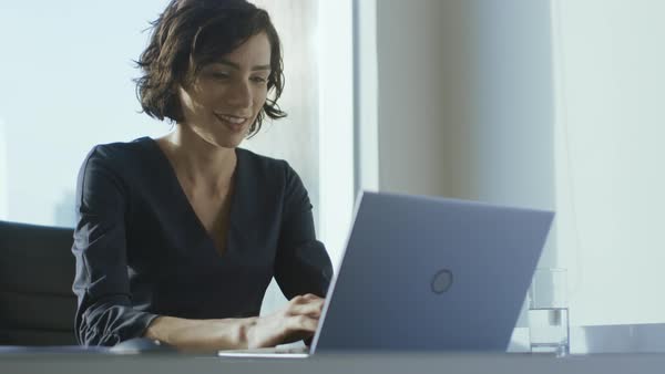 Confident female executive works on a laptop sitting at her desk in ...