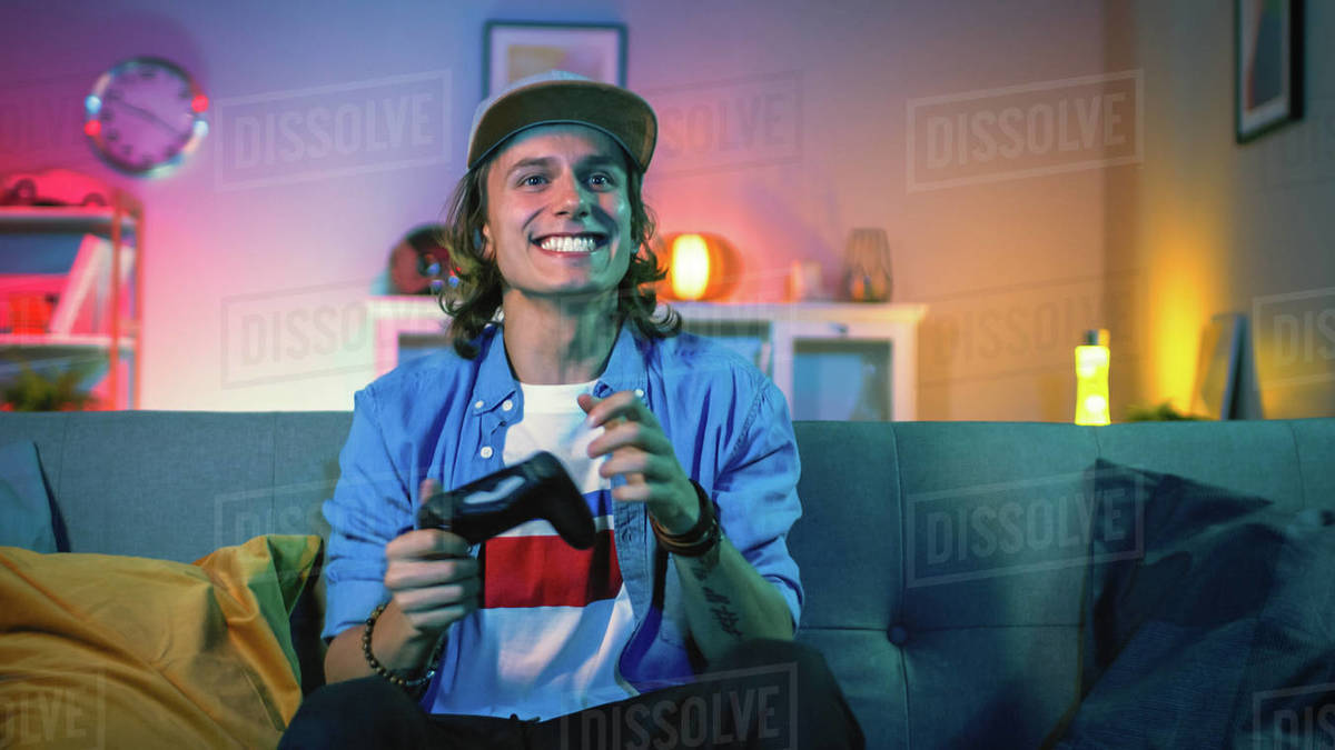 Handsome excited young gamer with long hair and a cap is sitting on a ...