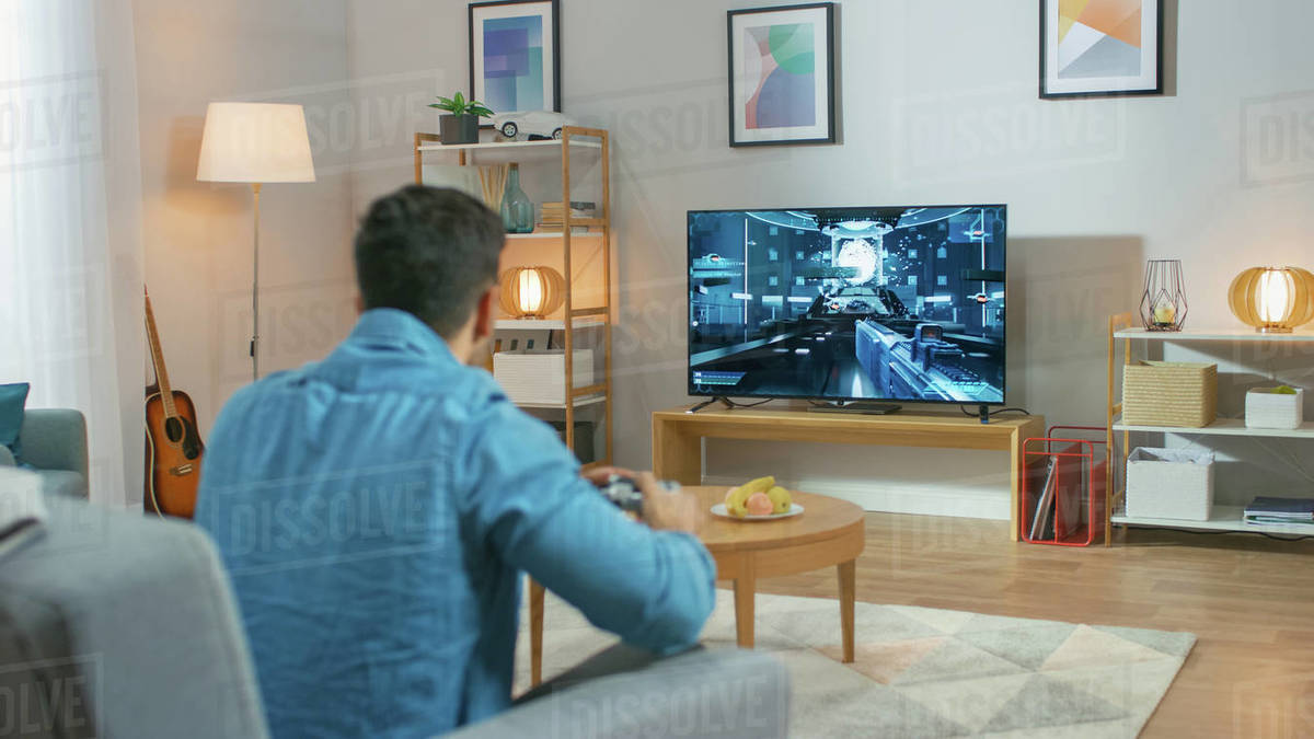 In the Living Room Man Sitting on a Couch Holds Controller Playing in a ...