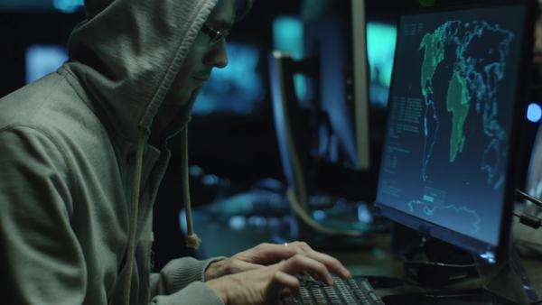 Two hackers in hoods work on a computers with maps and data on display ...