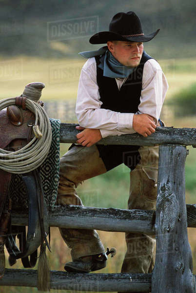 Agriculture - A young cowboy leans on a rustic corral rail / near ...
