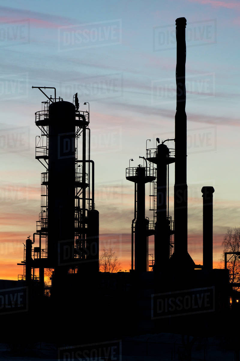 Silhouetted towers of a gas plant at sunset with orange cast clouds and ...