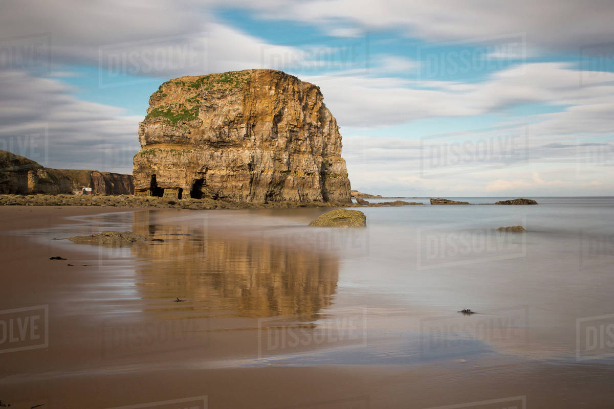 A large rock formation reflected in the wet sand on a beach on the ...