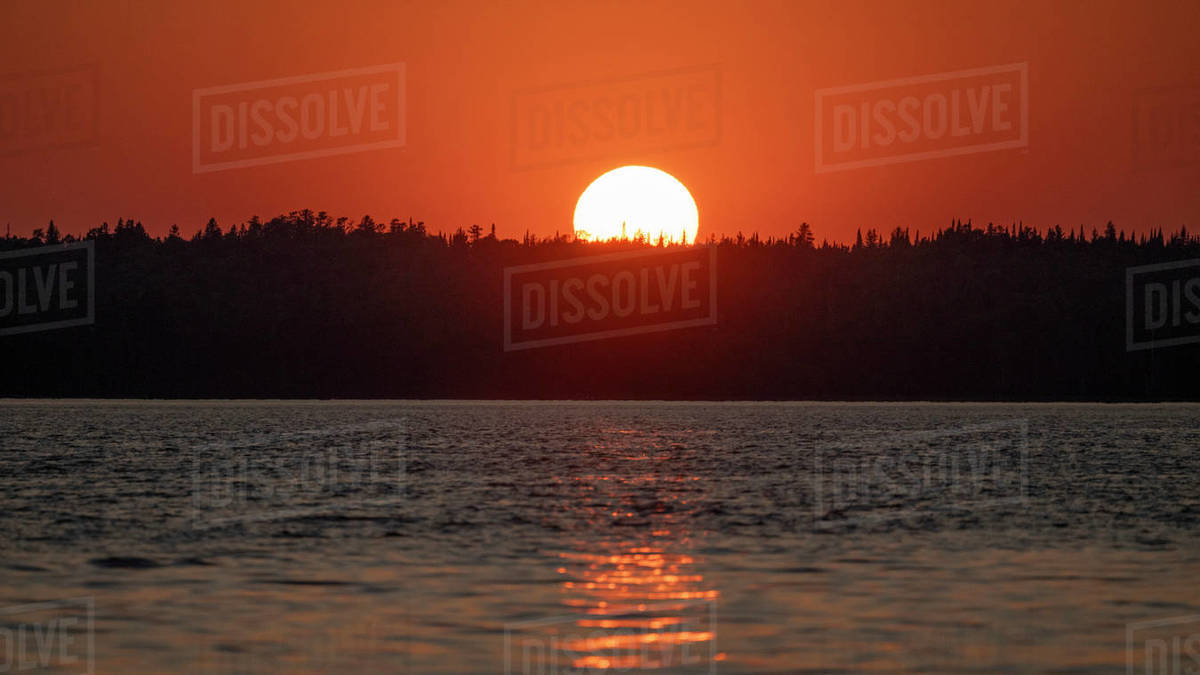 Glowing sun setting over a lake with silhouetted trees along the ...