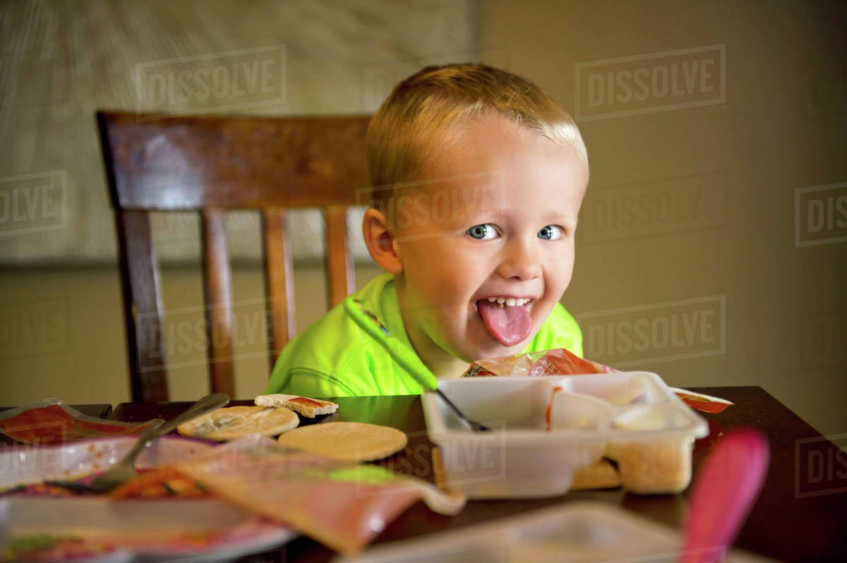 Young boy makes a silly face sticking out his tongue while eating his ...