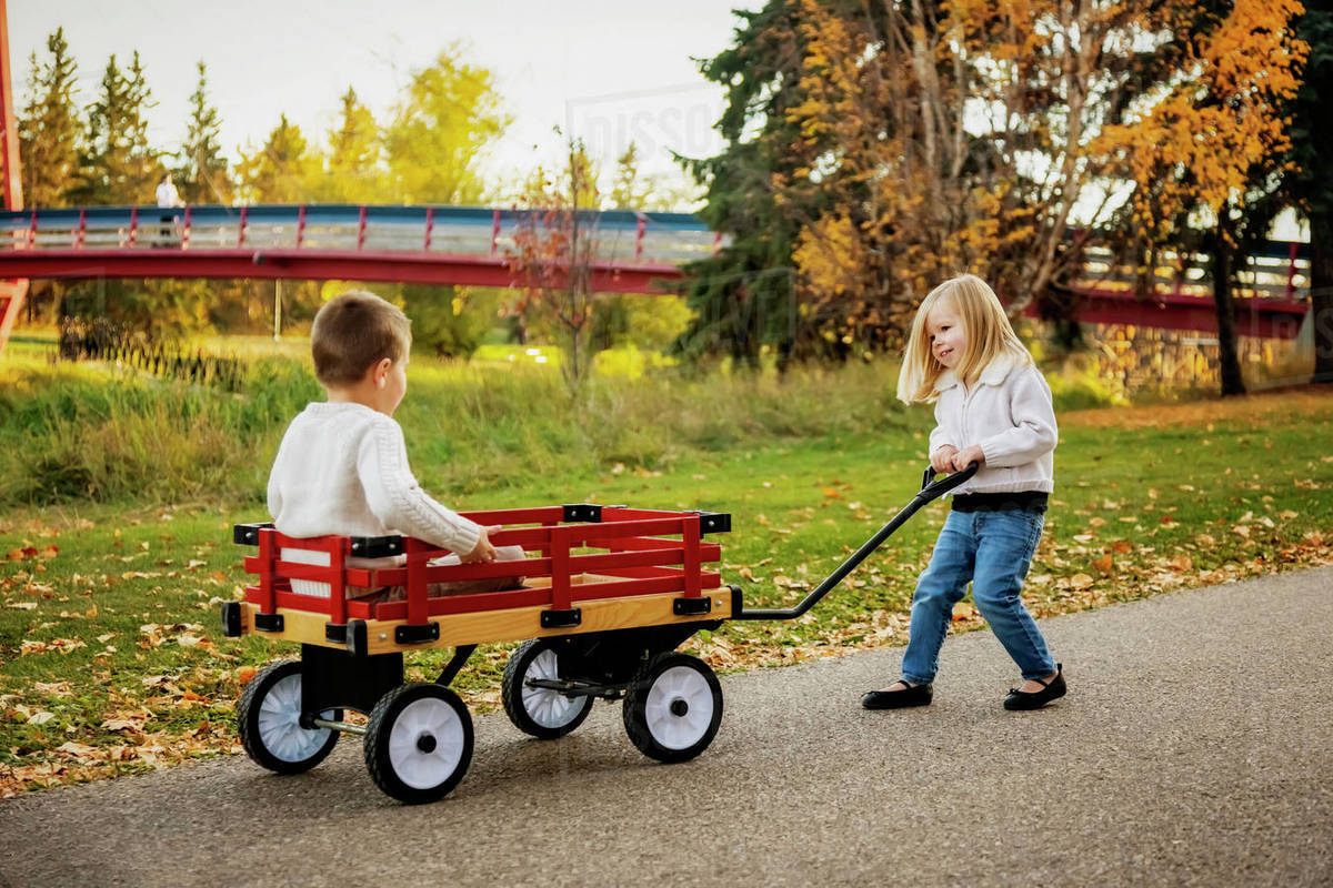 Young girl pulling her brother in a wagon at a city park along a river ...