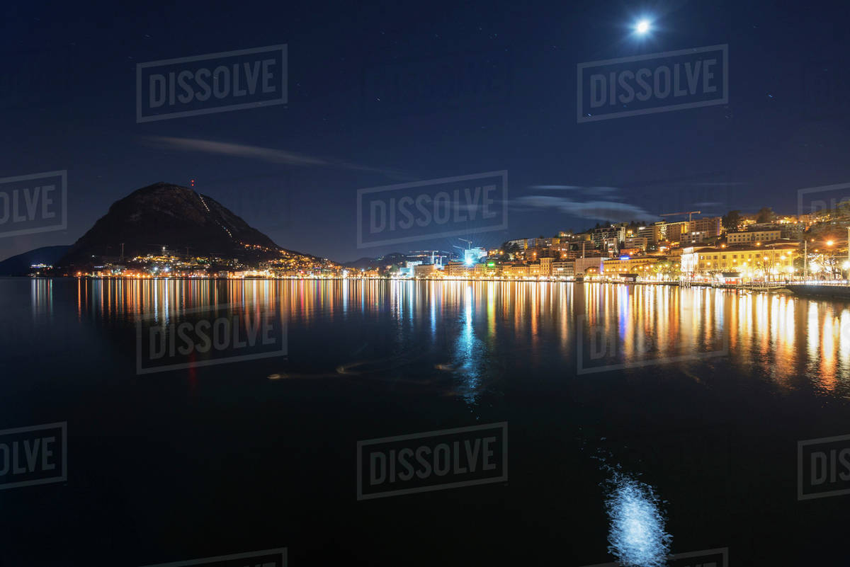 City lights illuminated on the tranquil Lake Lugano; Lugano, Ticino ...