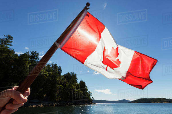 A man holds a Canadian Flag into the sun with the Gulf Islands the ...