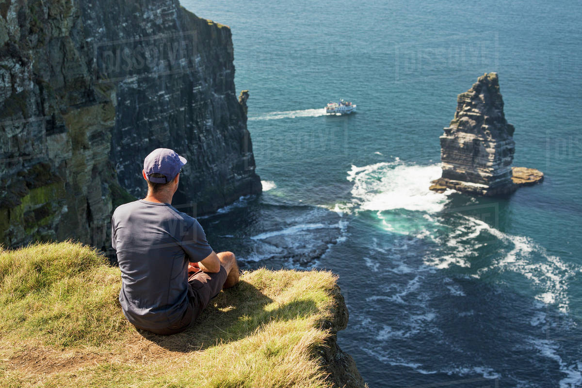 Man sitting on grassy cliff's edge overlooking large sea stack rock ...