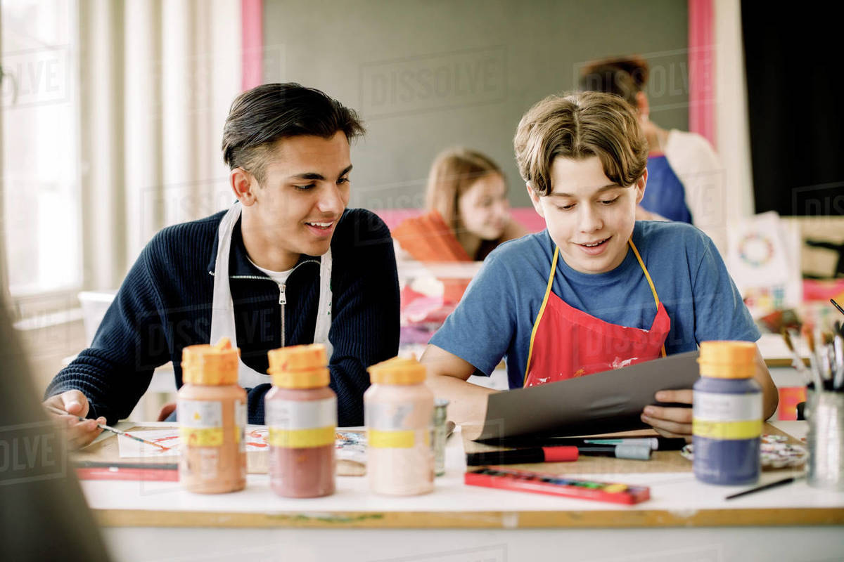 Smiling teenage boy showing artwork to male friend during painting ...