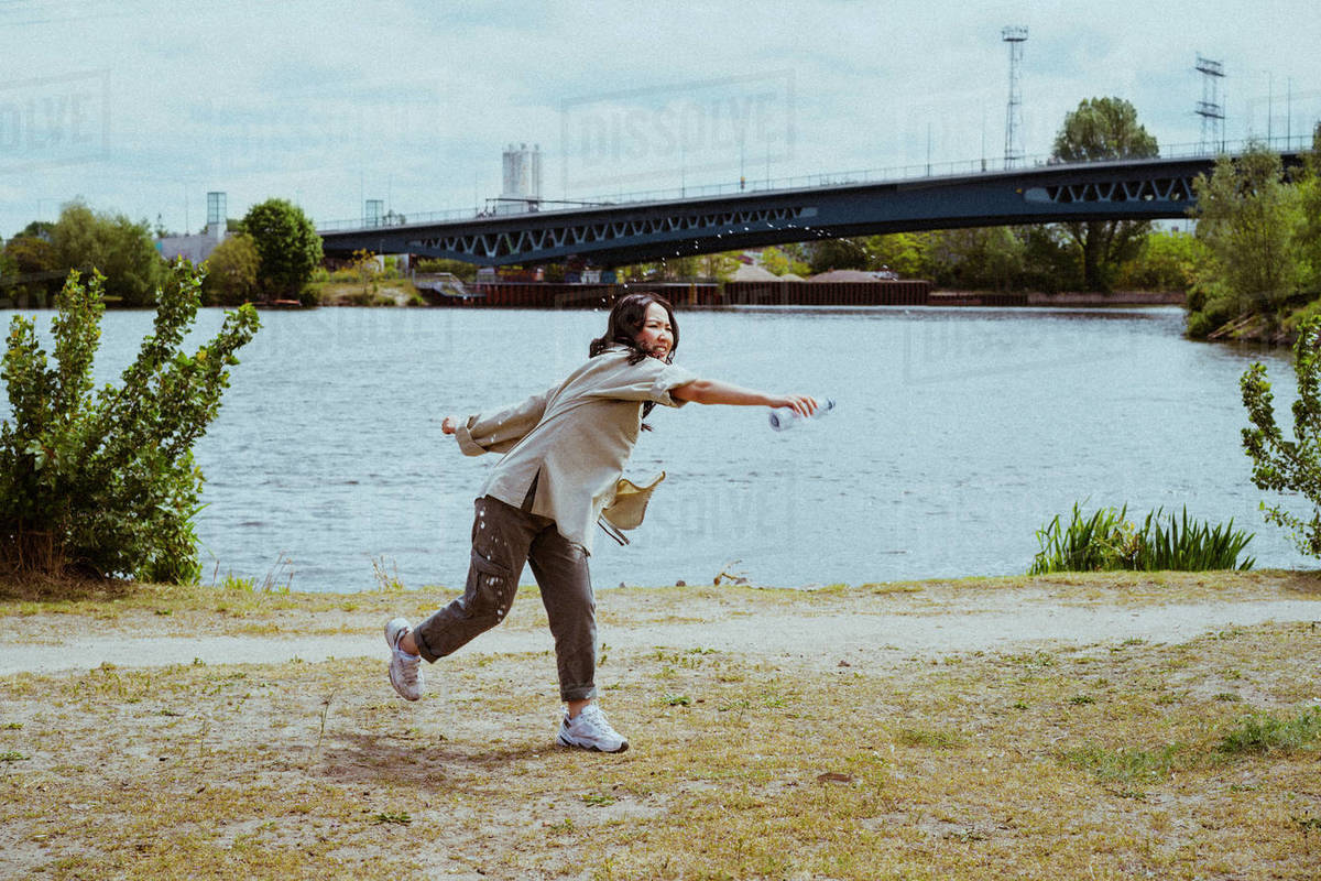 Full length of young woman throwing water from bottle near river ...