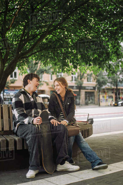 Happy male and female teenage friends laughing while sitting on bench ...