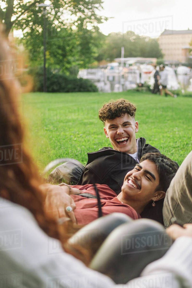 Happy male friends laughing together while talking with friend at park ...