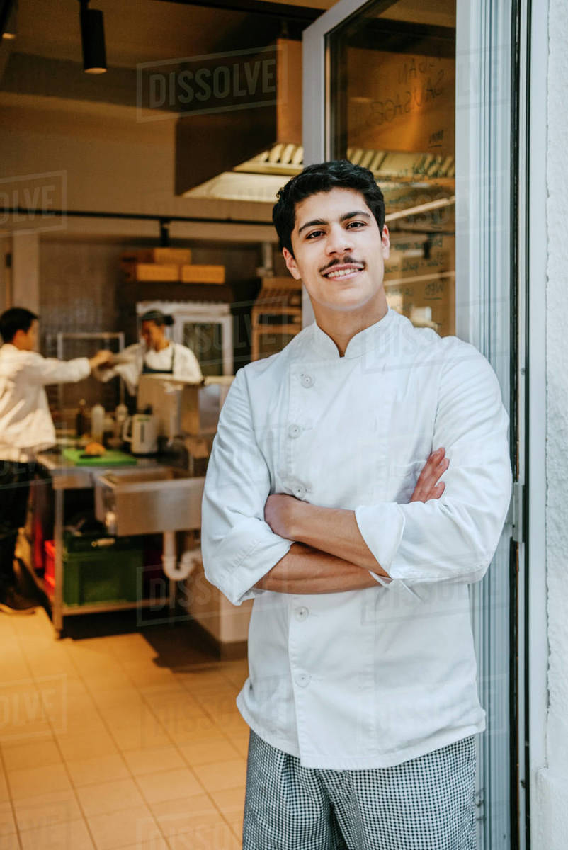 Portrait of happy male chef with arms crossed standing at door in ...
