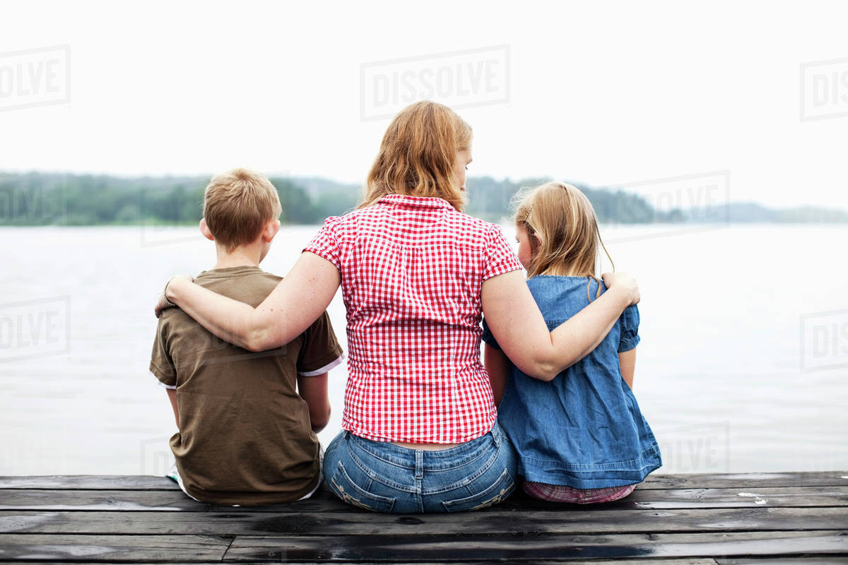 Rear view of mother with arm around her children sitting on pier ...