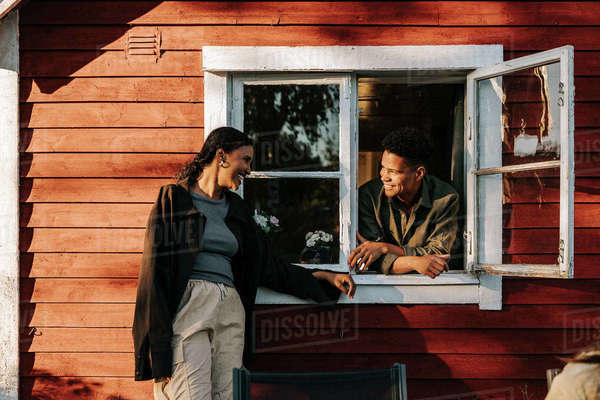 Smiling young man leaning on window and talking with female friend ...