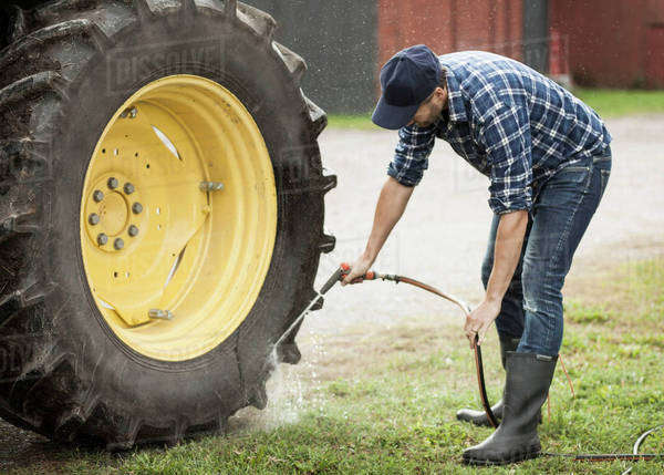 Full length of man washing tractor wheel with hose in farm - Royalty ...