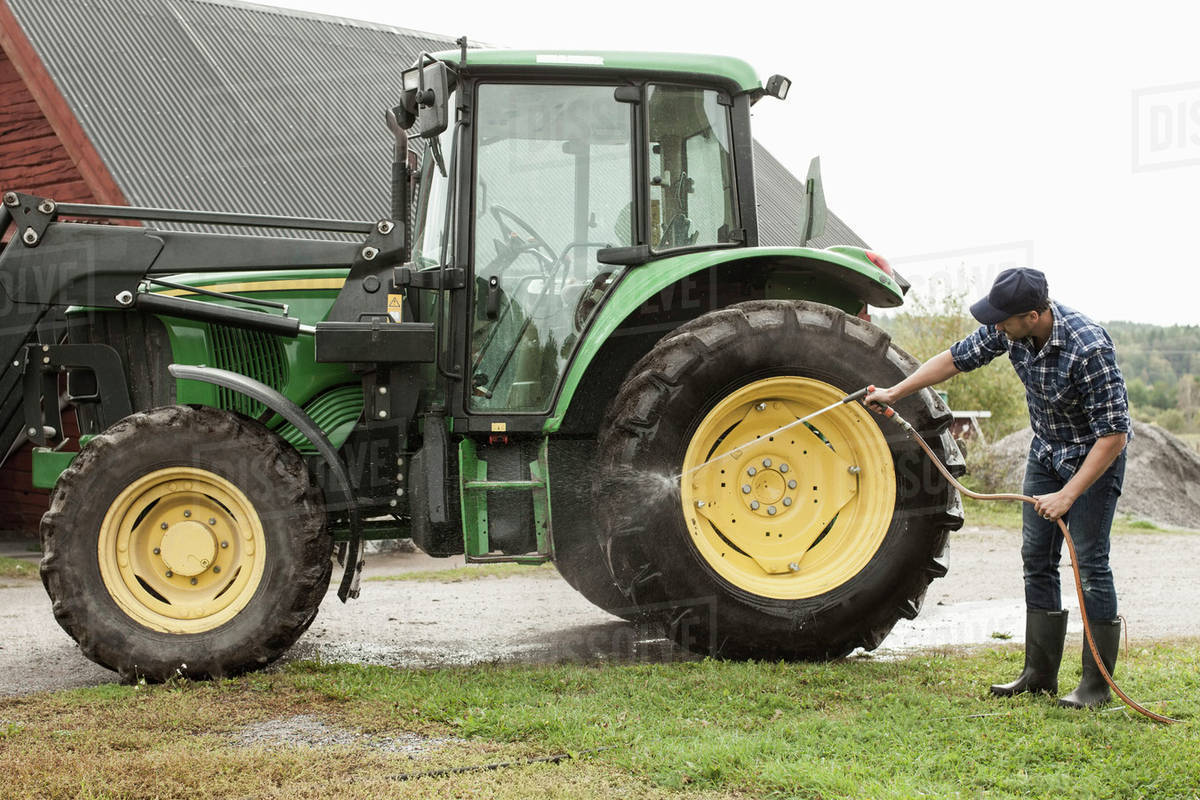 Full length farmer washing tractor wheel with hose in farm - Stock ...