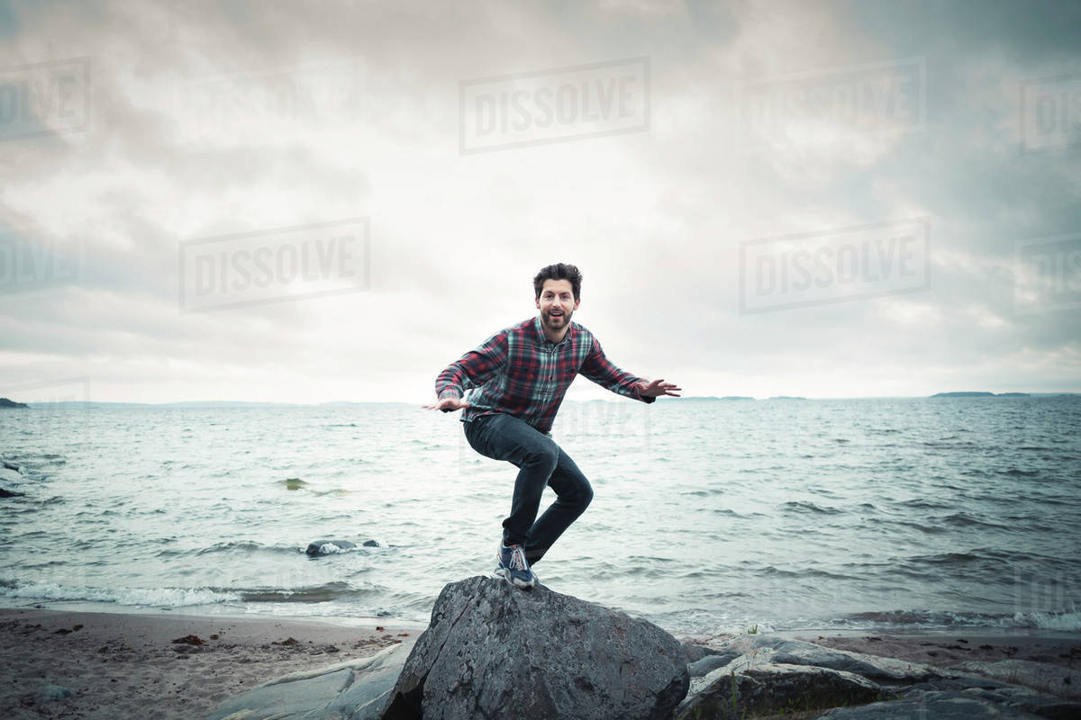 Full length portrait of happy man on rock by sea against cloudy sky ...
